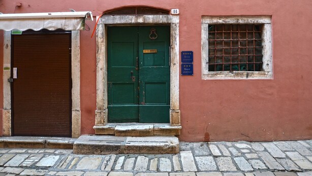 Um edifício com uma porta verde, uma janela com grade de metal e uma porta marrom fechada.