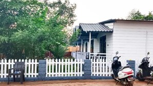 Uma casa com fachada branca, varanda e cerca branca de piquete. Duas scooters estão estacionadas na frente.