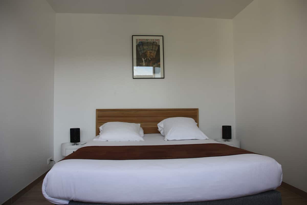 Cama bem arrumada com roupa de cama branca e castanha, cabeceira de madeira e duas mesas de cabeceira com alto-falantes.