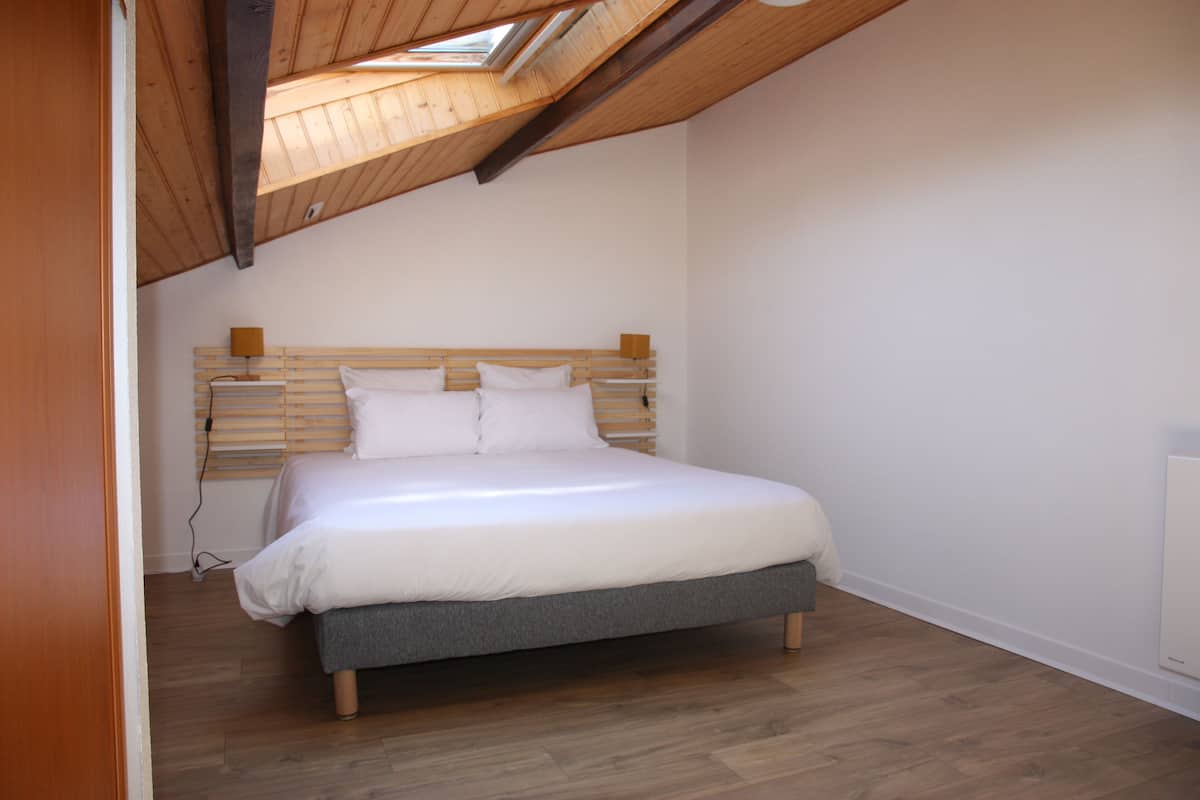 Quarto com teto inclinado, uma cama com cabeceira de madeira e aquecedor fixado na parede.
