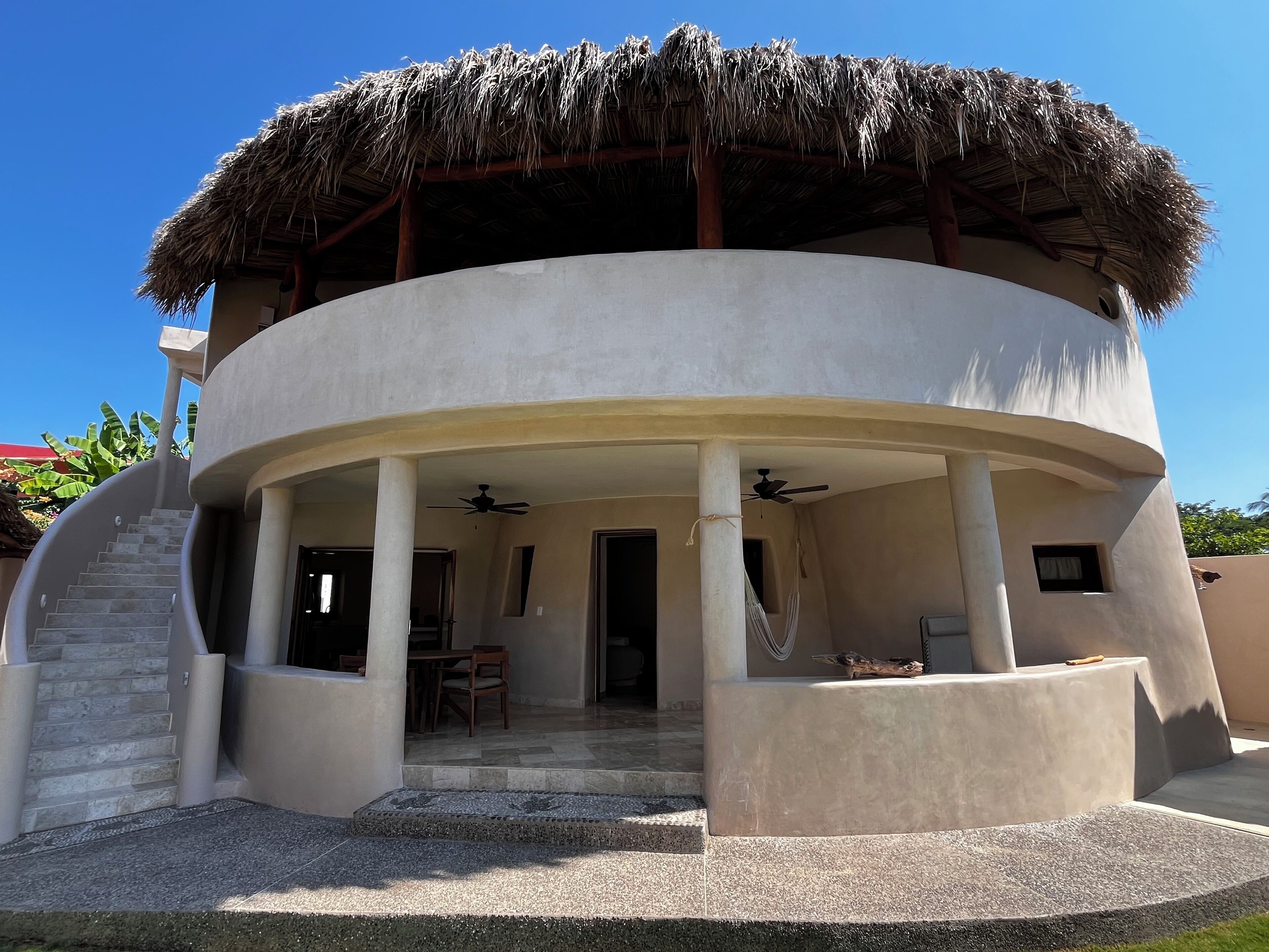 Um edifício moderno e circular com telhado de palha, uma área externa coberta com assentos e uma escadaria que leva à entrada.