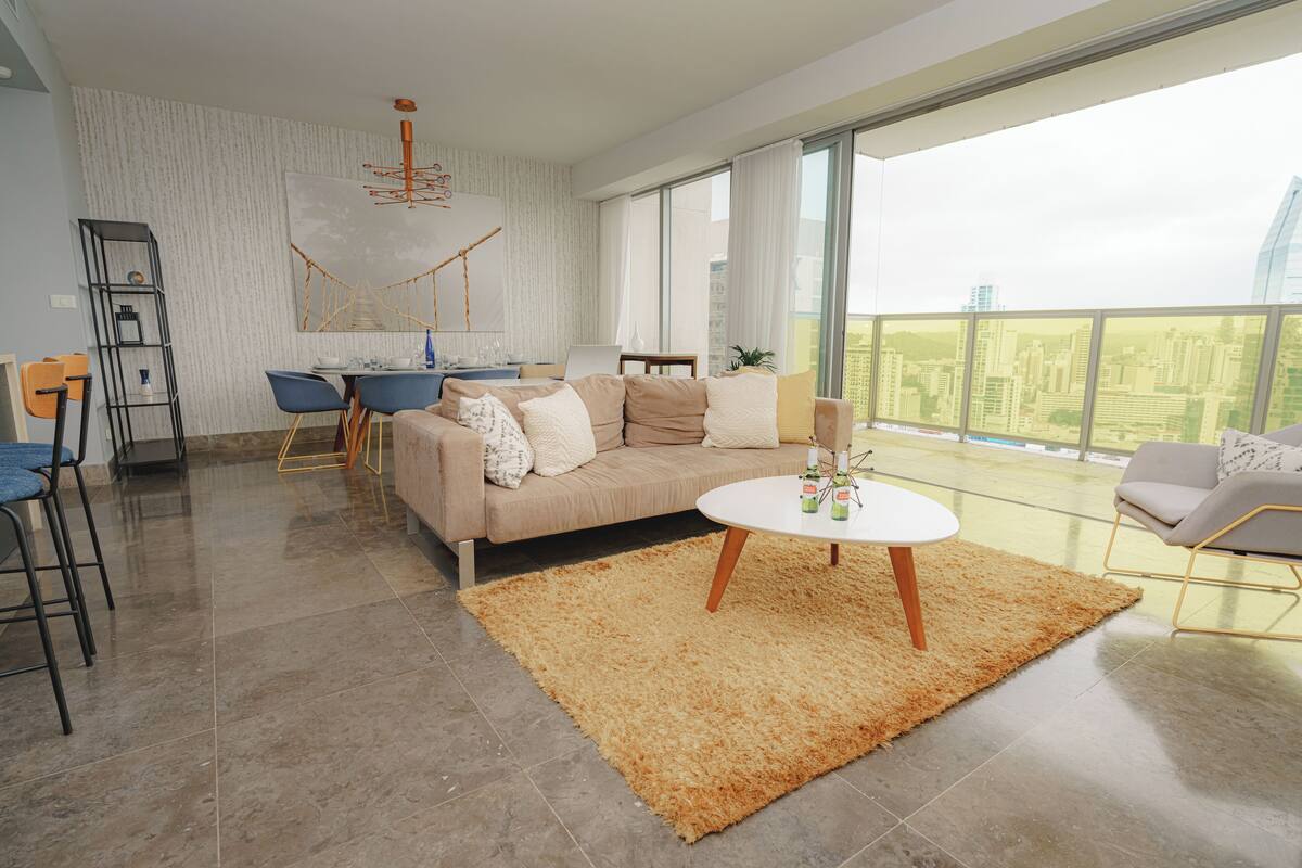 Uma sala de estar moderna com um sofá bege, uma mesa de centro redonda e vista para a cidade através de grandes janelas.