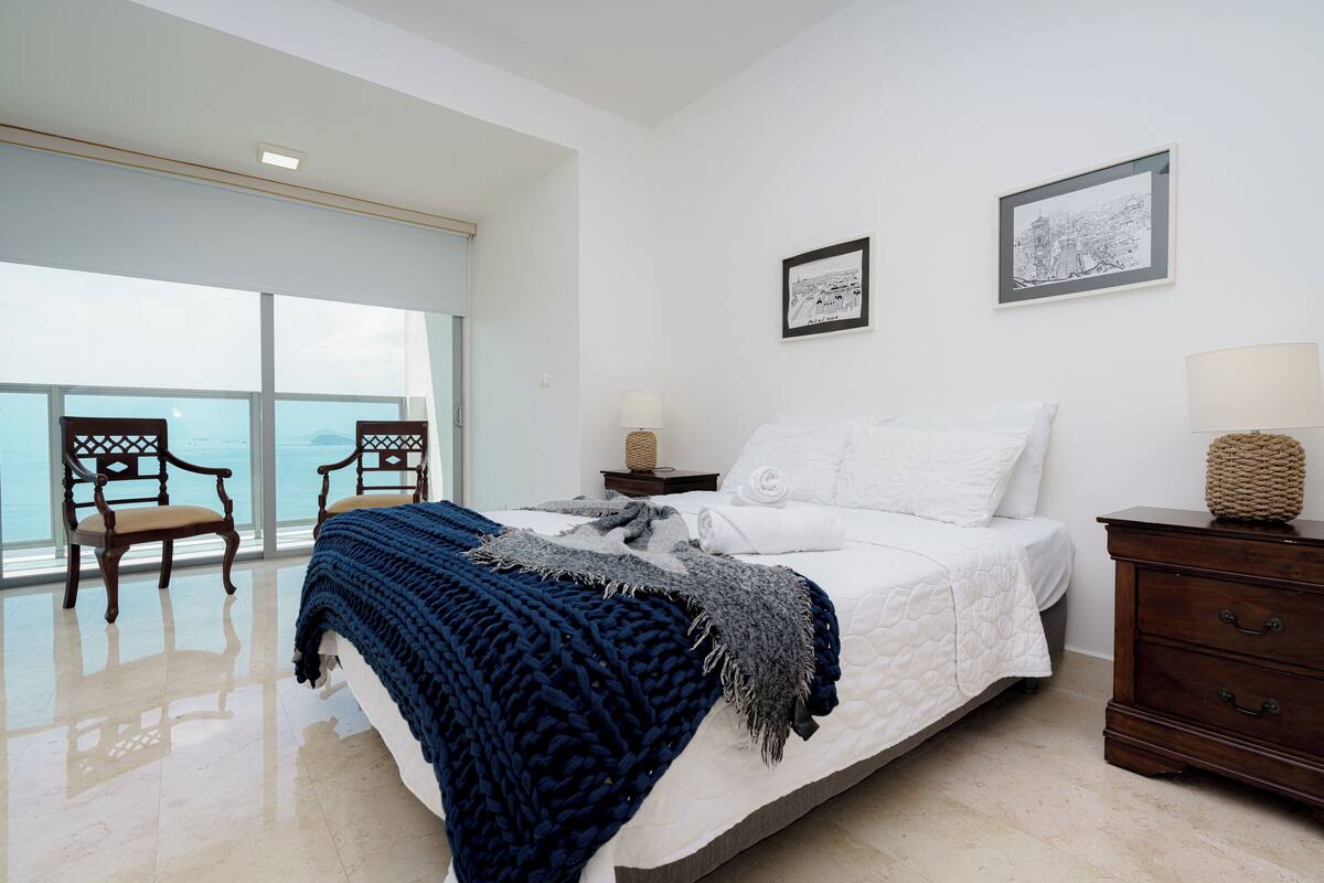 Quarto com uma cama grande, uma mesa de cabeceira, cômoda, duas cadeiras e vista para o mar através de grandes janelas.