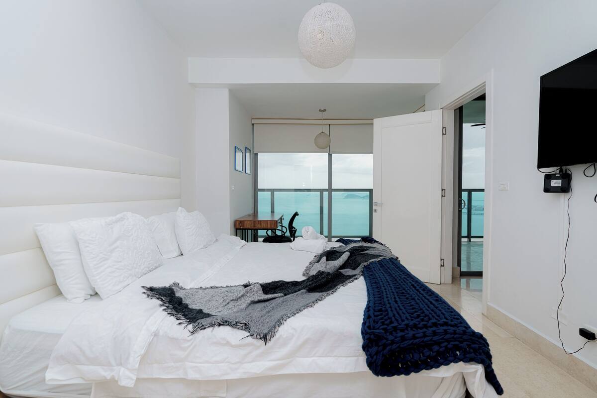 Quarto moderno com uma cama grande, vista para o mar através de uma porta de vidro e uma televisão na parede.