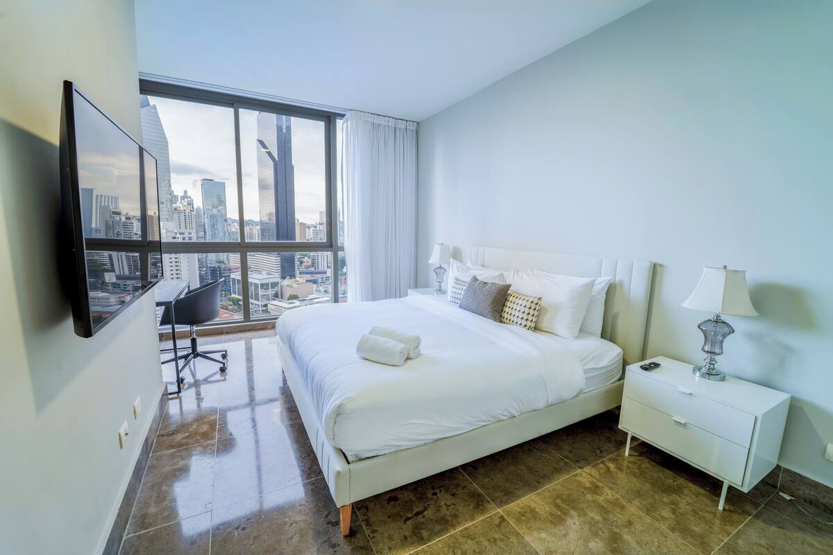 Quarto de hotel moderno com uma cama grande, uma escrivaninha, uma cadeira e vista para o horizonte da cidade através de amplas janelas.