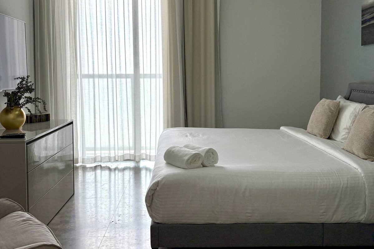 Quarto moderno com uma cama grande, mesa de cabeceira, luminária e janela com cortinas.