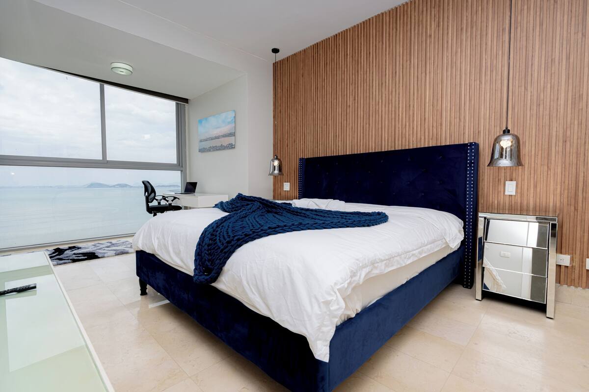Quarto moderno com uma cama grande, vista para o mar e painel de parede de madeira.