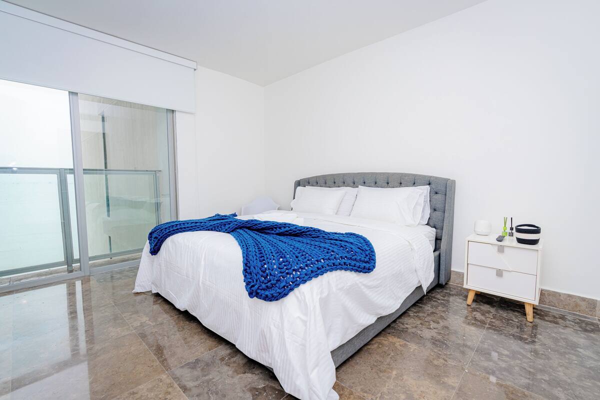 Quarto moderno com uma cama grande, uma mesa de cabeceira e uma porta de vidro que dá para uma varanda.