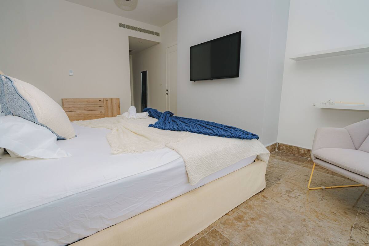Quarto com cama, televisão na parede, cabeceira de madeira e uma cadeira branca.