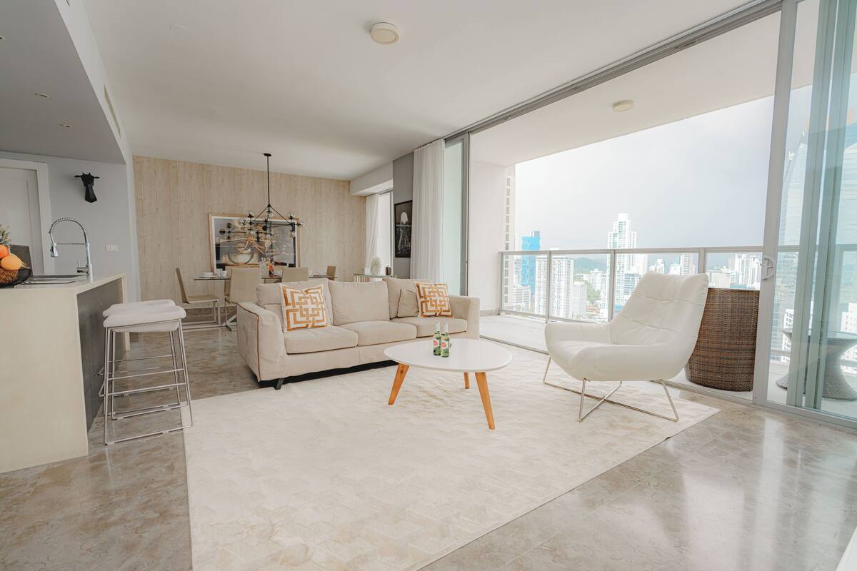 Uma sala moderna com sofá, mesa de centro e uma poltrona, uma área de jantar com mesa e cadeiras, e uma grande janela com vista para a cidade.