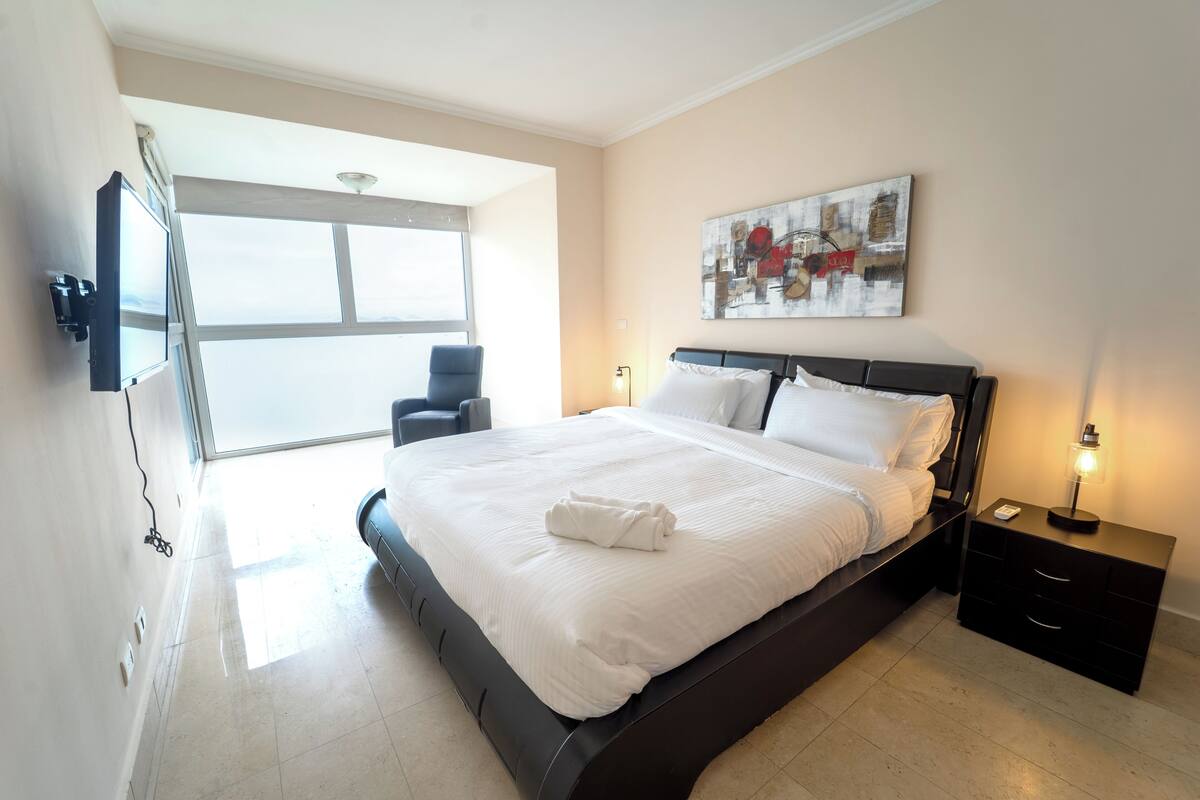 Quarto de hotel moderno com uma cama grande, televisão de tela plana na parede, mesa de cabeceira com luminária e janela com vista.