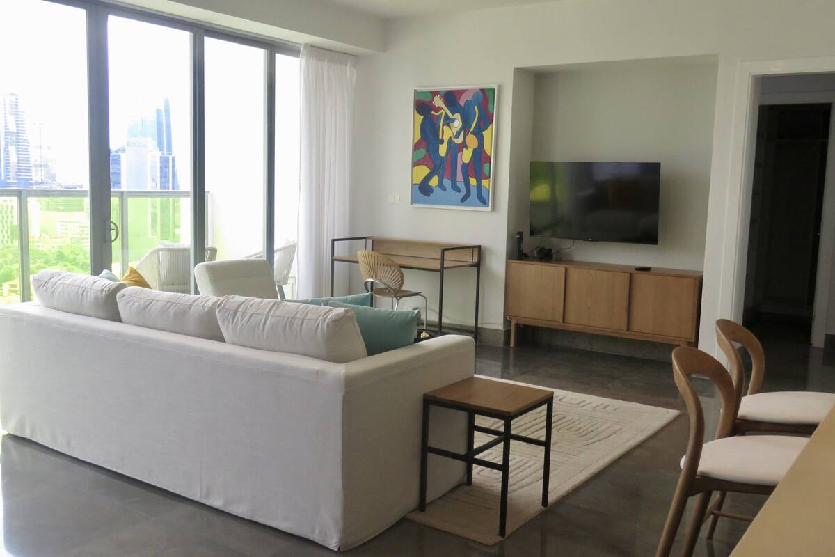 Sala moderna com sofá branco, televisão e vista para a cidade.