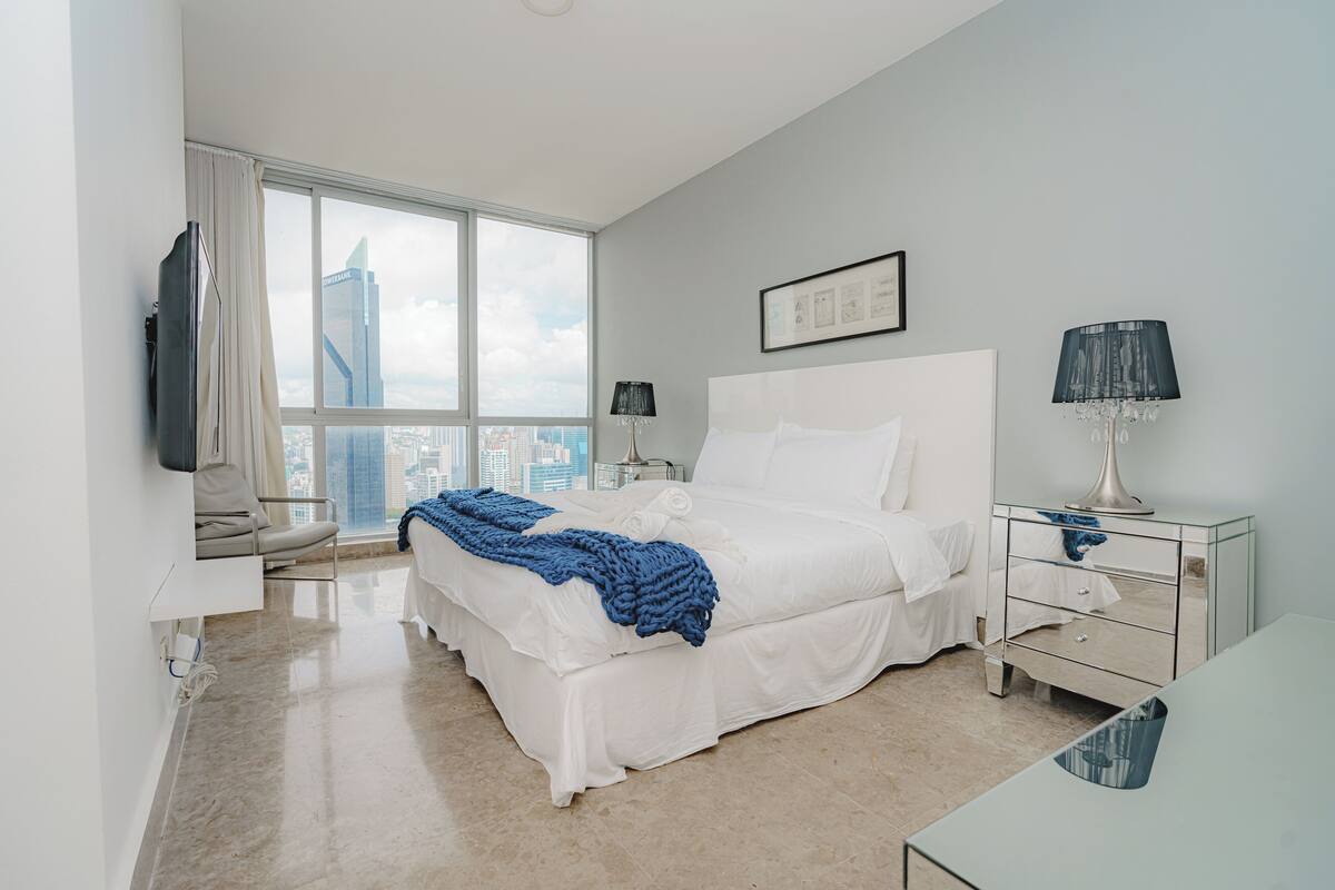 Quarto de hotel moderno com uma cama grande, uma mesa de cabeceira com abajur, uma cadeira e vista para o horizonte da cidade.