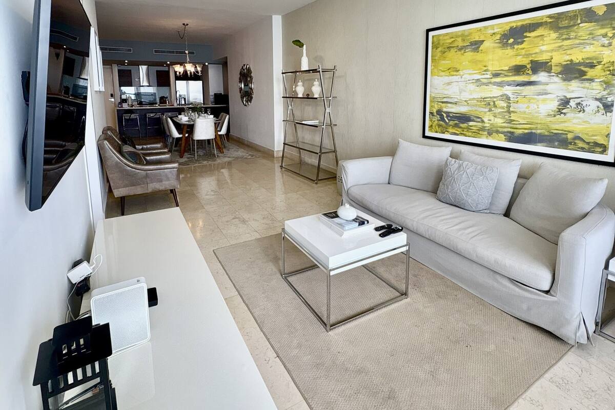 Uma sala de estar moderna com um sofá branco, mesa de centro e TV fixada na parede. Ao fundo, há uma área de jantar com mesa e cadeiras.