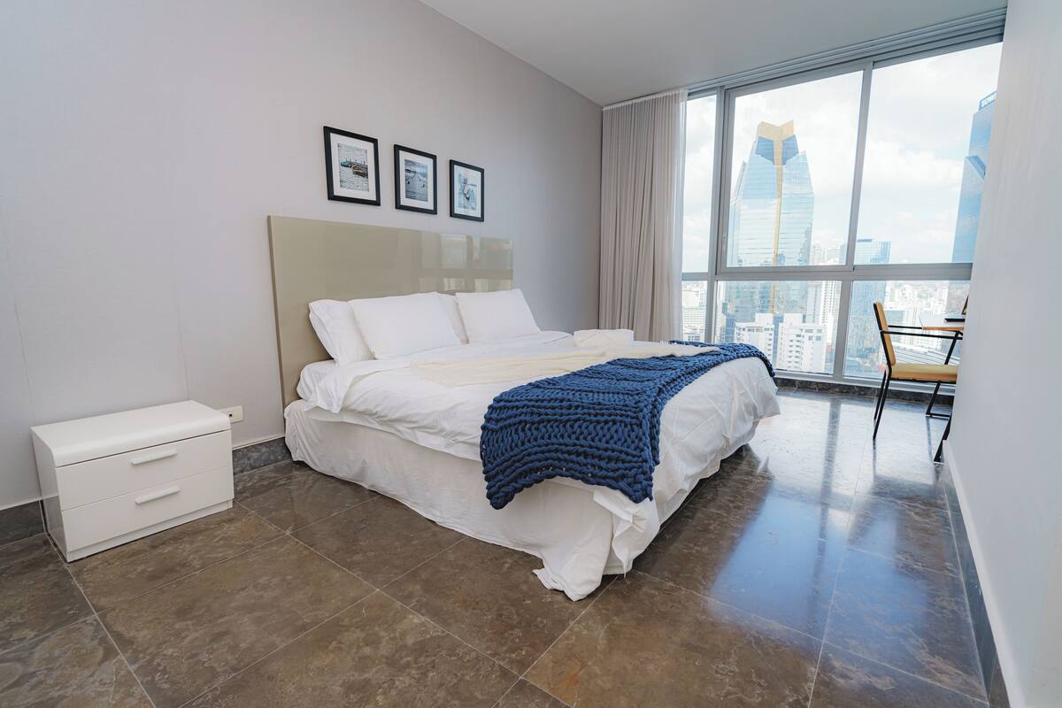 Quarto de hotel moderno com cama grande, mesa de cabeceira, cadeira e vista para a cidade pela janela.