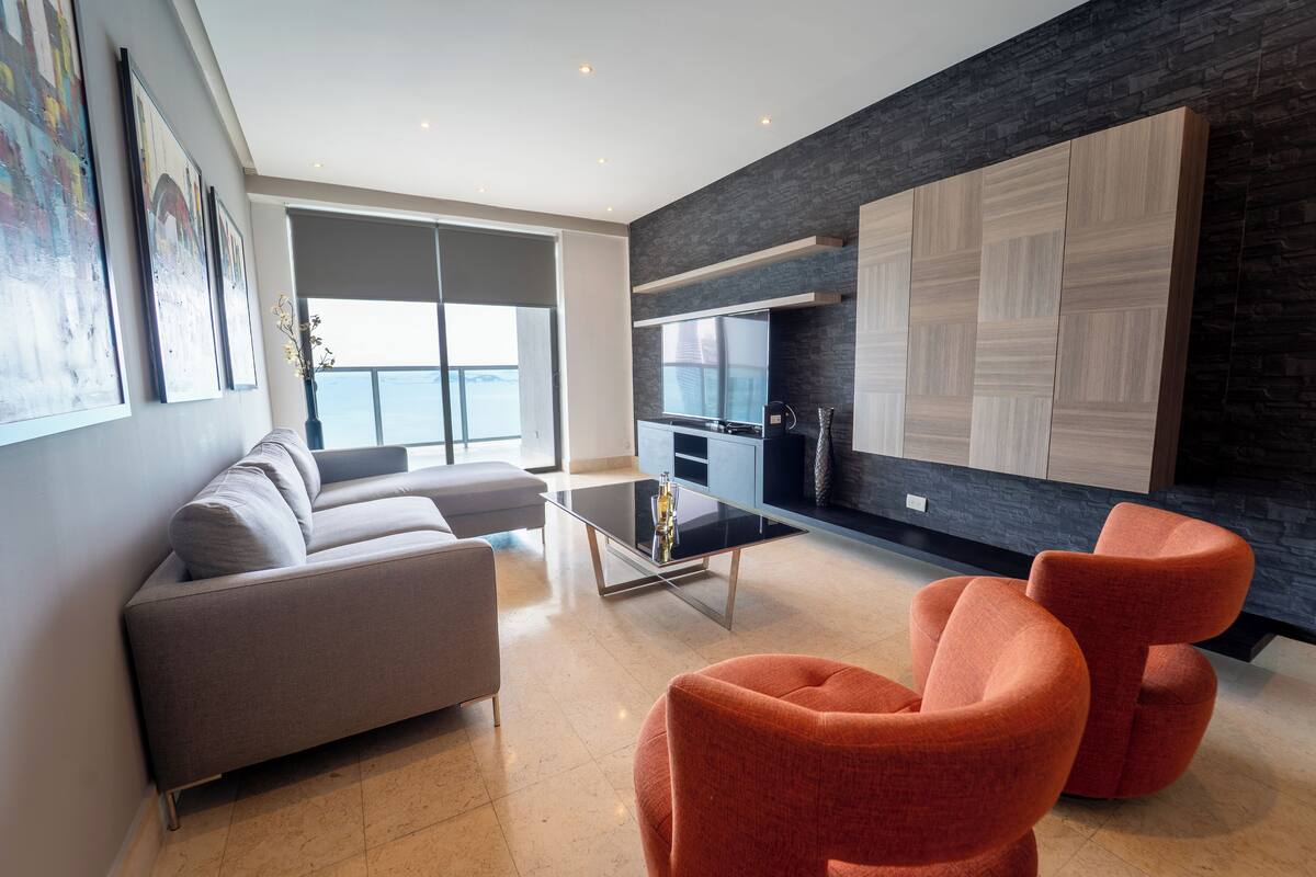 Uma sala de estar moderna com um sofá cinza, poltronas laranja, uma mesa de centro de vidro e uma televisão grande.