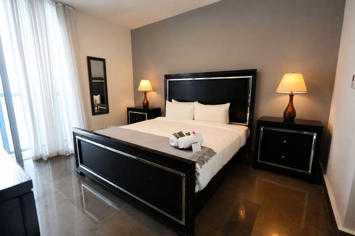 Quarto de hotel com uma cama grande, duas mesas de cabeceira com luminárias, um espelho e vista para o exterior.