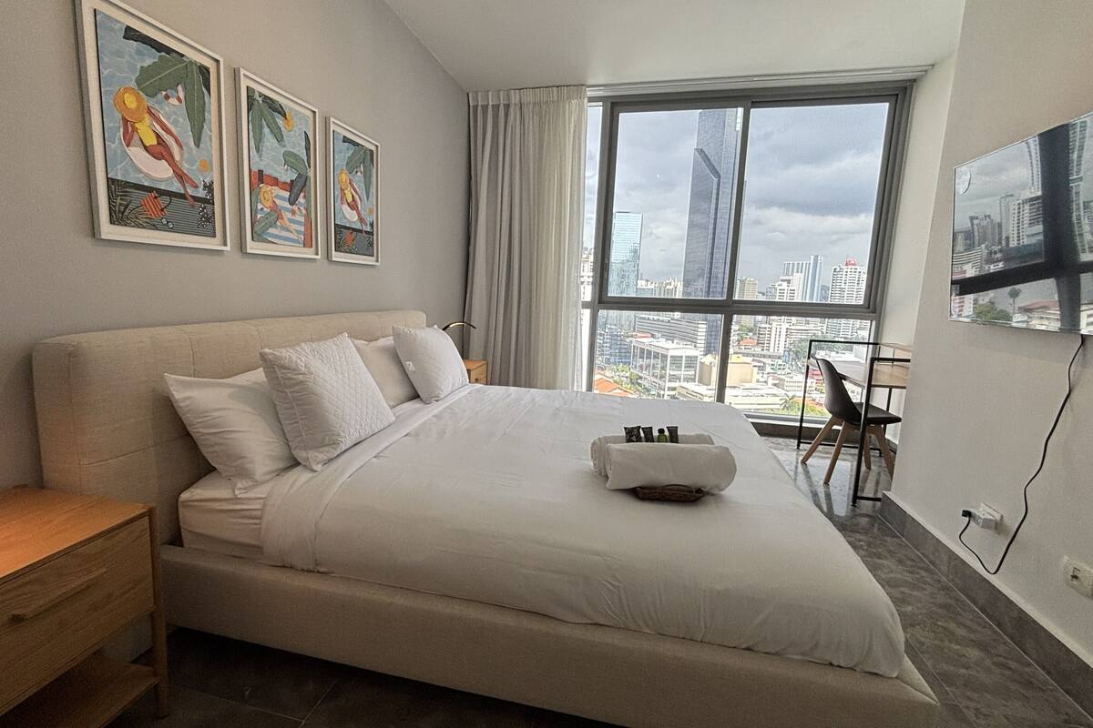 Quarto de hotel moderno com uma cama grande, uma mesa de cabeceira, uma escrivaninha e vista para a cidade pela janela.