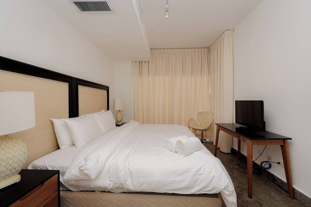 Quarto de hotel com cama, escrivaninha, cadeira, televisão e abajur.