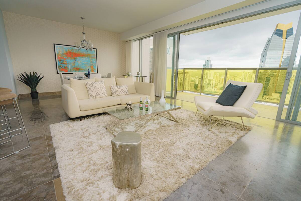 Uma sala de estar moderna com um sofá branco, uma mesa de centro de vidro e um tapete felpudo branco. Há uma janela grande com vista para um horizonte urbano e um edifício.