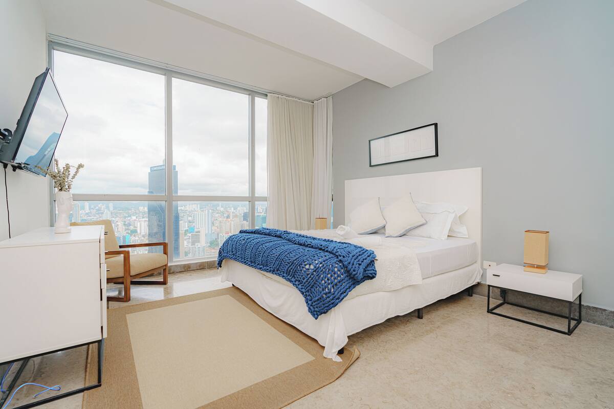Quarto moderno com uma janela grande, uma cama, uma mesinha de cabeceira, uma cadeira e uma escrivaninha.