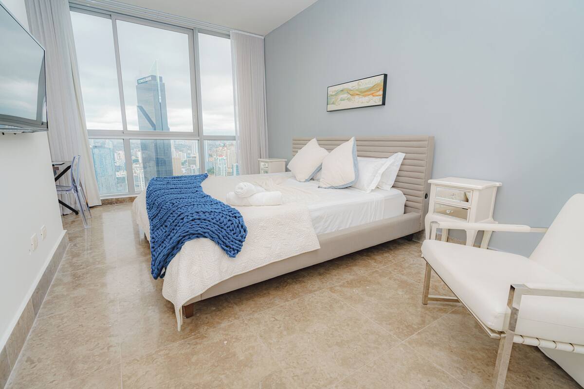 Quarto de hotel moderno com uma cama grande, mesa de cabeceira, cadeira e vista para a cidade pela janela.