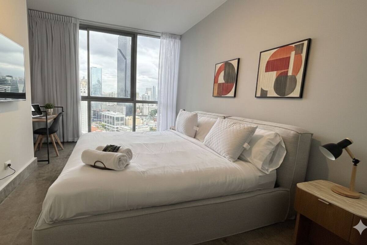 Quarto de hotel moderno com uma cama grande, uma escrivaninha com um laptop, uma TV e vista para a cidade pela janela.
