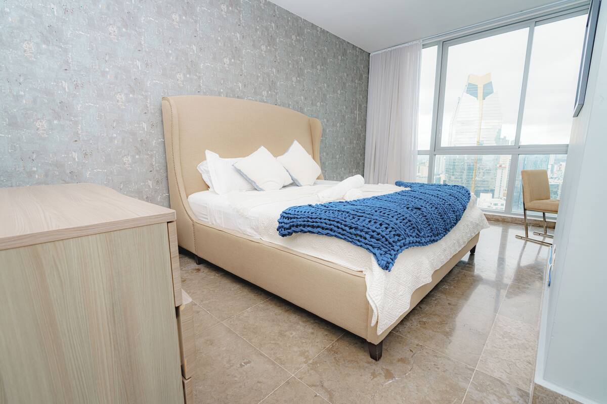 Quarto moderno com uma cama grande, uma mesinha de cabeceira e vista para a cidade através de uma janela ampla.