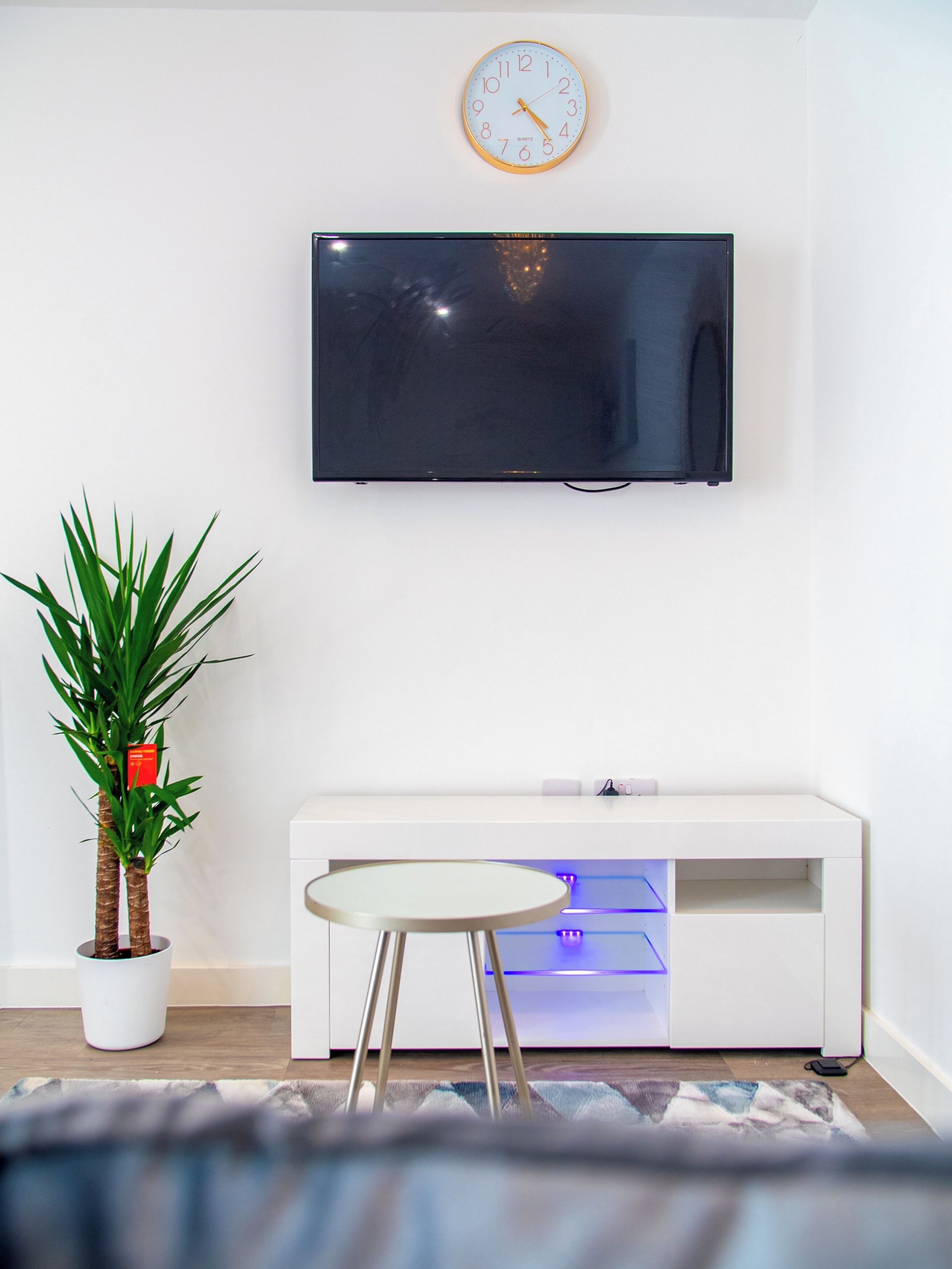 Uma sala de estar moderna com uma televisão de tela plana na parede, uma mesa redonda pequena, um móvel branco para eletrônicos com iluminação LED azul e um vaso de planta.