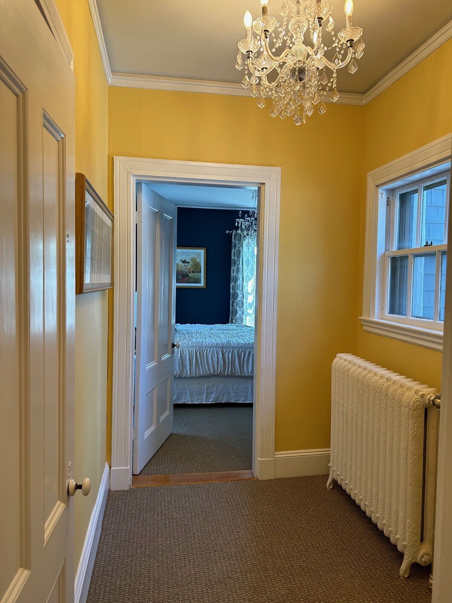 Um corredor que dá para um quarto com cama, radiador e um candelabro.