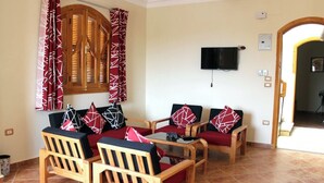 Uma sala de estar com móveis de madeira, almofadas vermelhas, uma televisão e uma janela com cortinas.