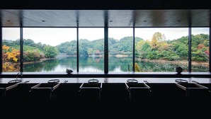 Quarto com grande janela com vista para um lago e floresta. Há várias mesas com cadeiras, uma pia e um vaso no parapeito da janela.