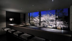 Um bar moderno com um balcão extenso, banquetas e vista para cerejeiras à noite.