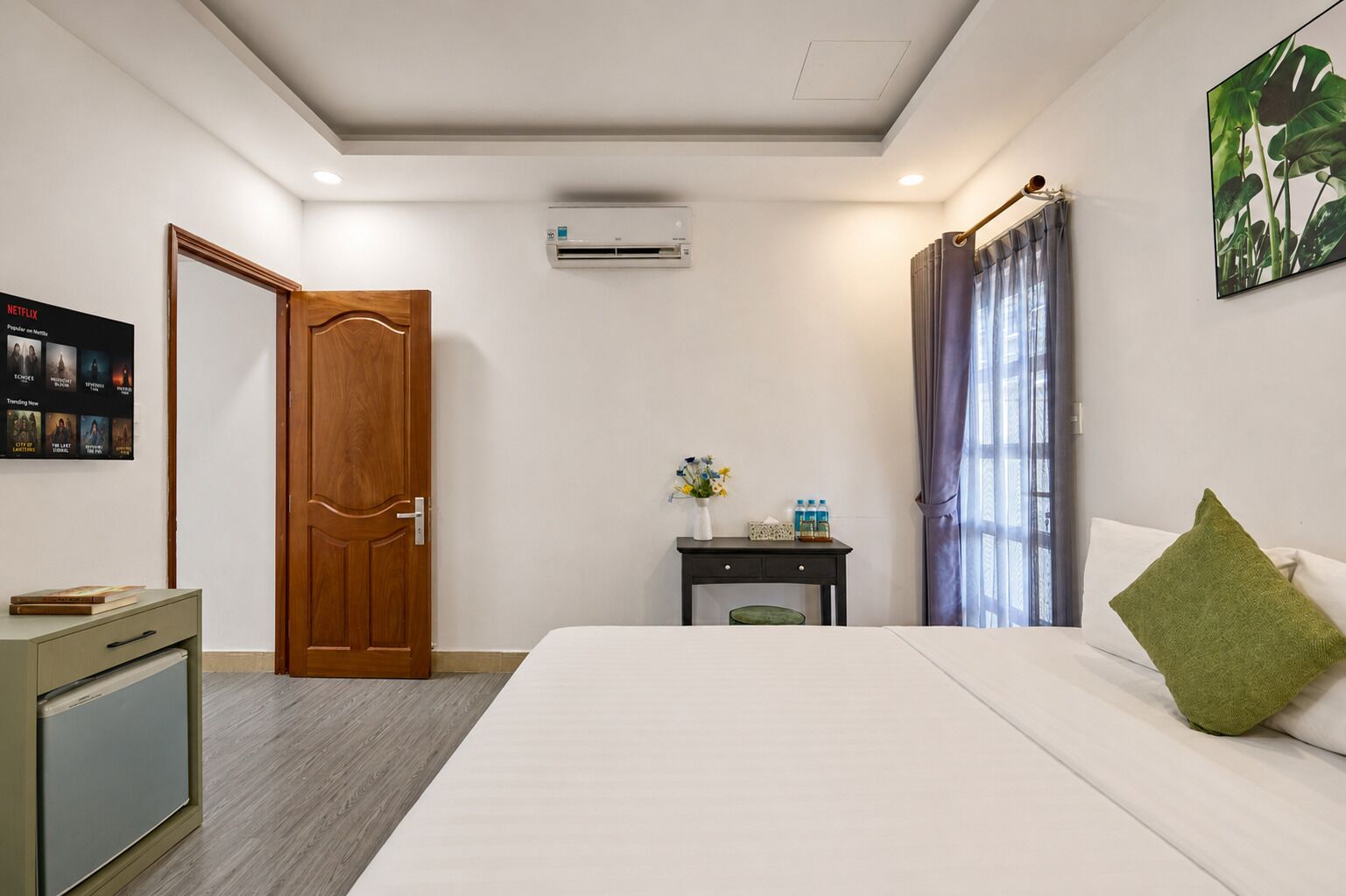 Quarto de hotel com cama, escrivaninha, guarda-roupa, ar condicionado, uma pequena mesa com um vaso e garrafas, e janela com cortinas.