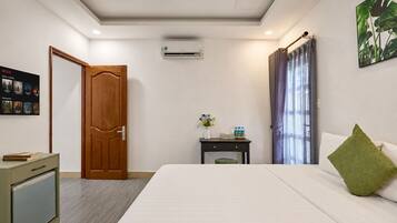 Quarto de hotel com cama, escrivaninha, guarda-roupa, ar condicionado, uma pequena mesa com um vaso e garrafas, e janela com cortinas.