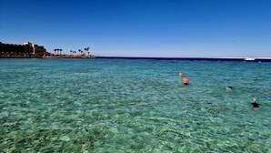 Mar azul cristalina com pessoas nadando e um prédio de resort ao fundo.