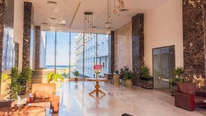 Um hall amplo com teto alto, grandes janelas com vista para a cidade e mobiliário moderno, incluindo poltronas de couro e uma mesa central.