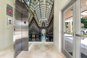 Corredor interno com teto de vidro, elevador e porta que dá para o exterior.