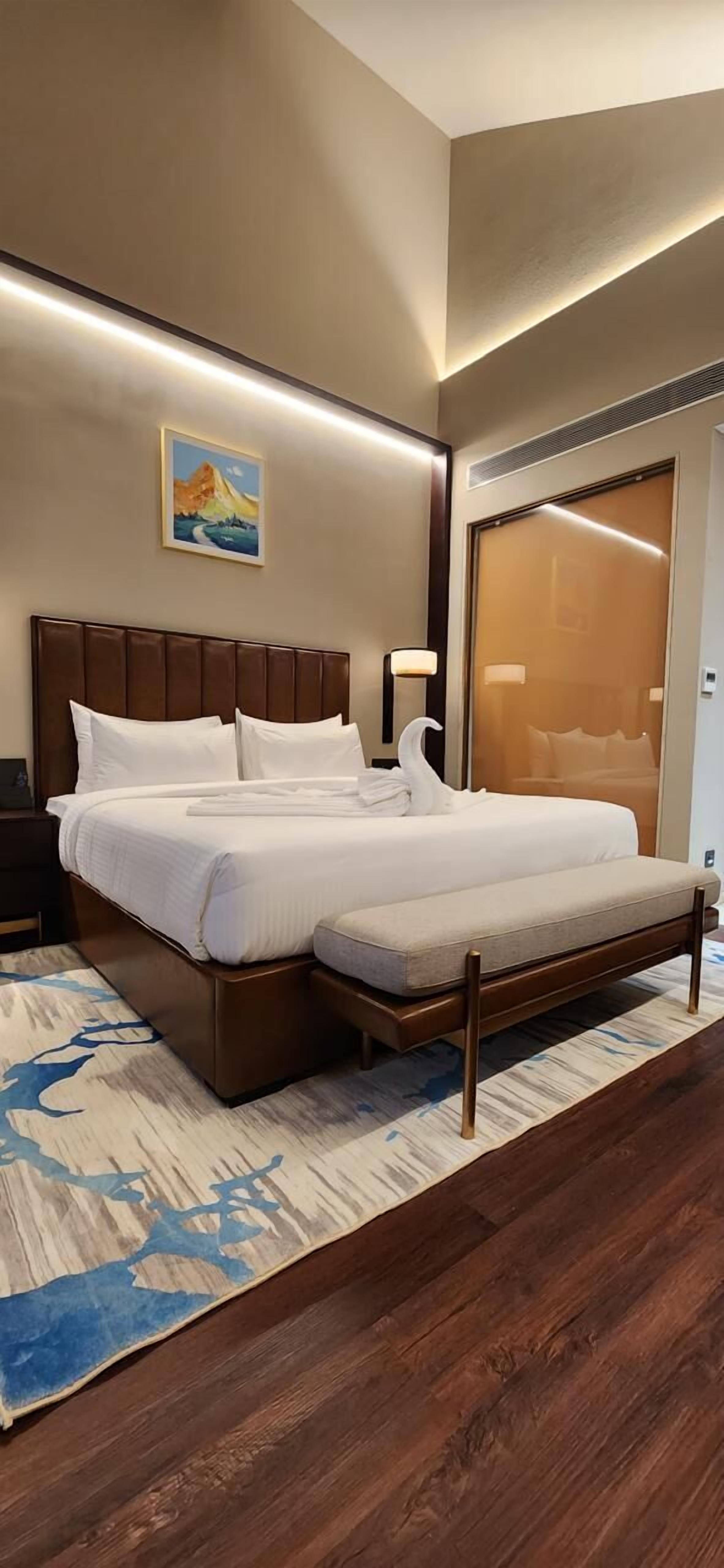 Quarto de hotel moderno com uma cama grande, uma mesa de cabeceira, uma luminária e um quadro na parede.