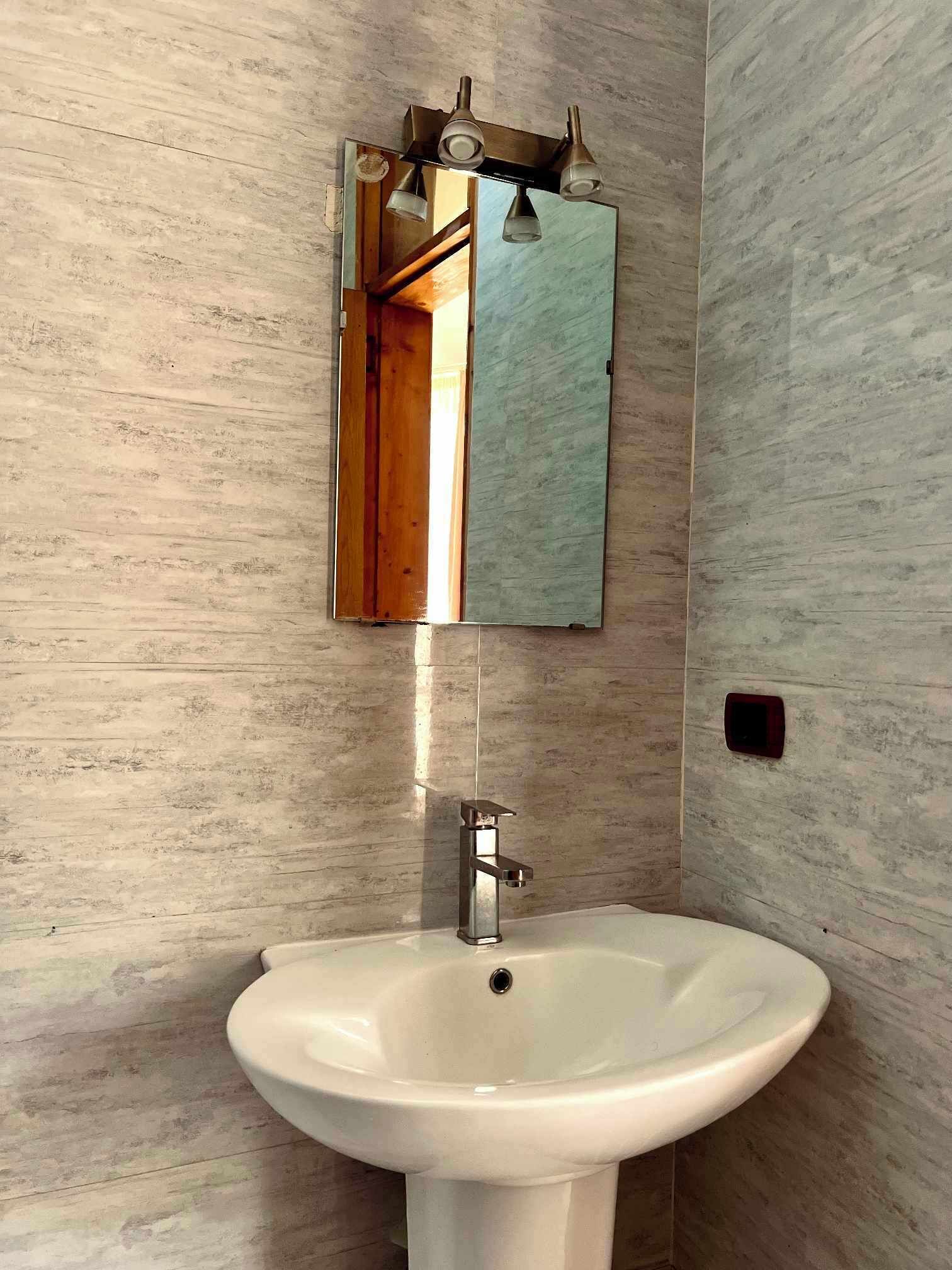 Casa de banho com lavatório branco, espelho e candeeiro fixado na parede.