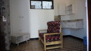 Quarto pequeno e compacto, com uma cadeira de madeira, um armário pequeno e uma janela com grades de metal.