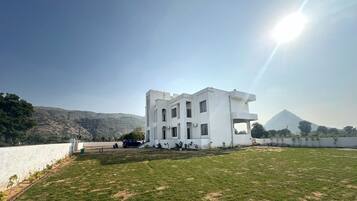 Um edifício moderno e branco, com telhado plano e várias janelas, rodeado por um relvado bem cuidado e uma montanha ao fundo.