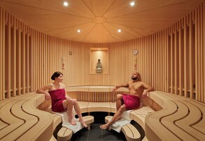 Uma sauna com bancos de madeira dispostos em semicírculo, uma estátua central e um relógio fixado na parede.