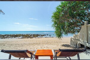Vista da praia com uma mesa de madeira e duas cadeiras, uma árvore e uma parede branca.