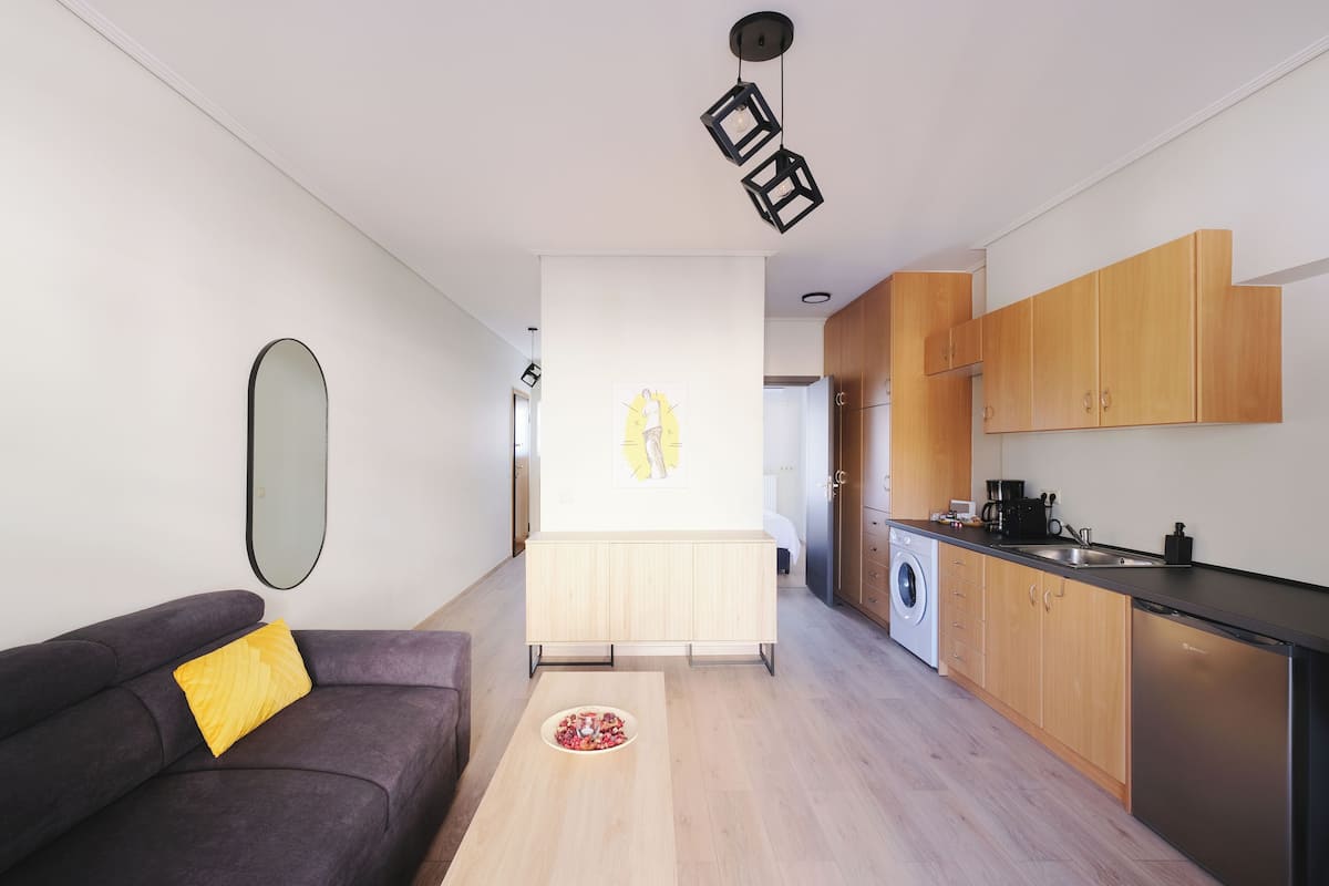 Um apartamento moderno com sofá, uma pequena mesa de jantar, cozinha com armários de madeira e máquina de lavar.