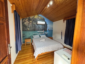 Quarto com teto de madeira, cama com colcha florida, mesa de cabeceira de madeira e eletrodoméstico branco. Na parede, um mural retrata uma paisagem costeira com um barco.