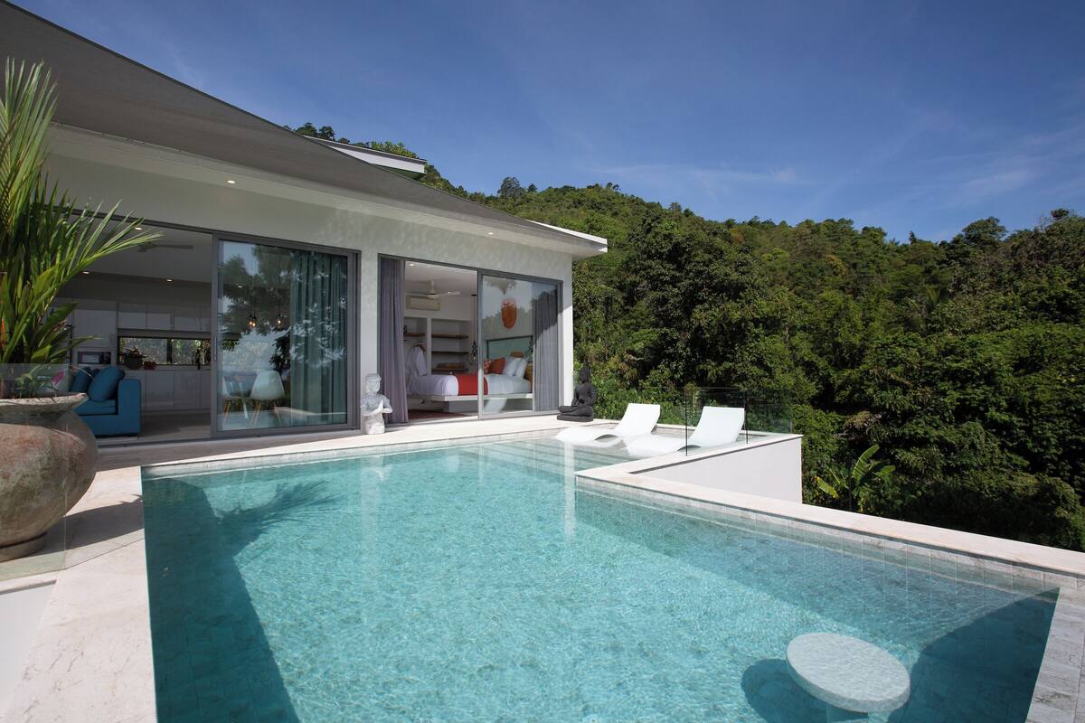 Uma casa moderna com piscina, amplas janelas de vidro e vista para uma encosta arborizada.
