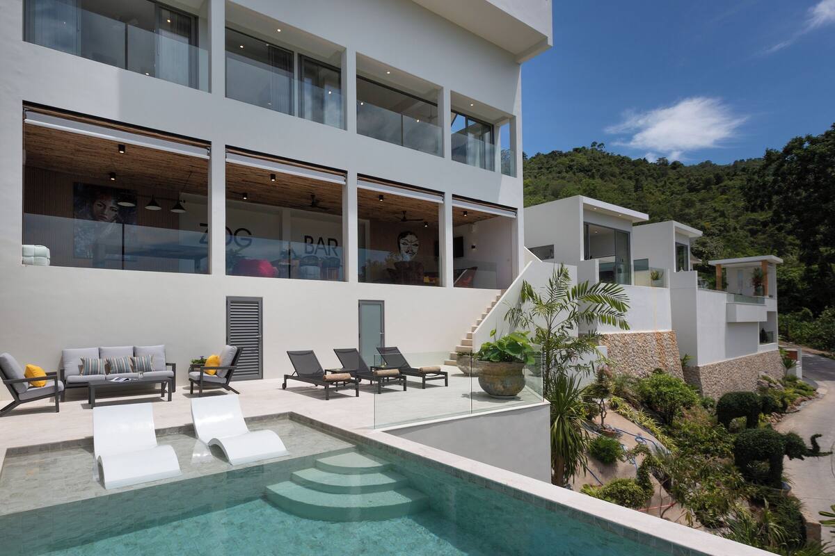 Um edifício moderno de dois andares com piscina no terraço, cadeiras de descanso e um bar.
