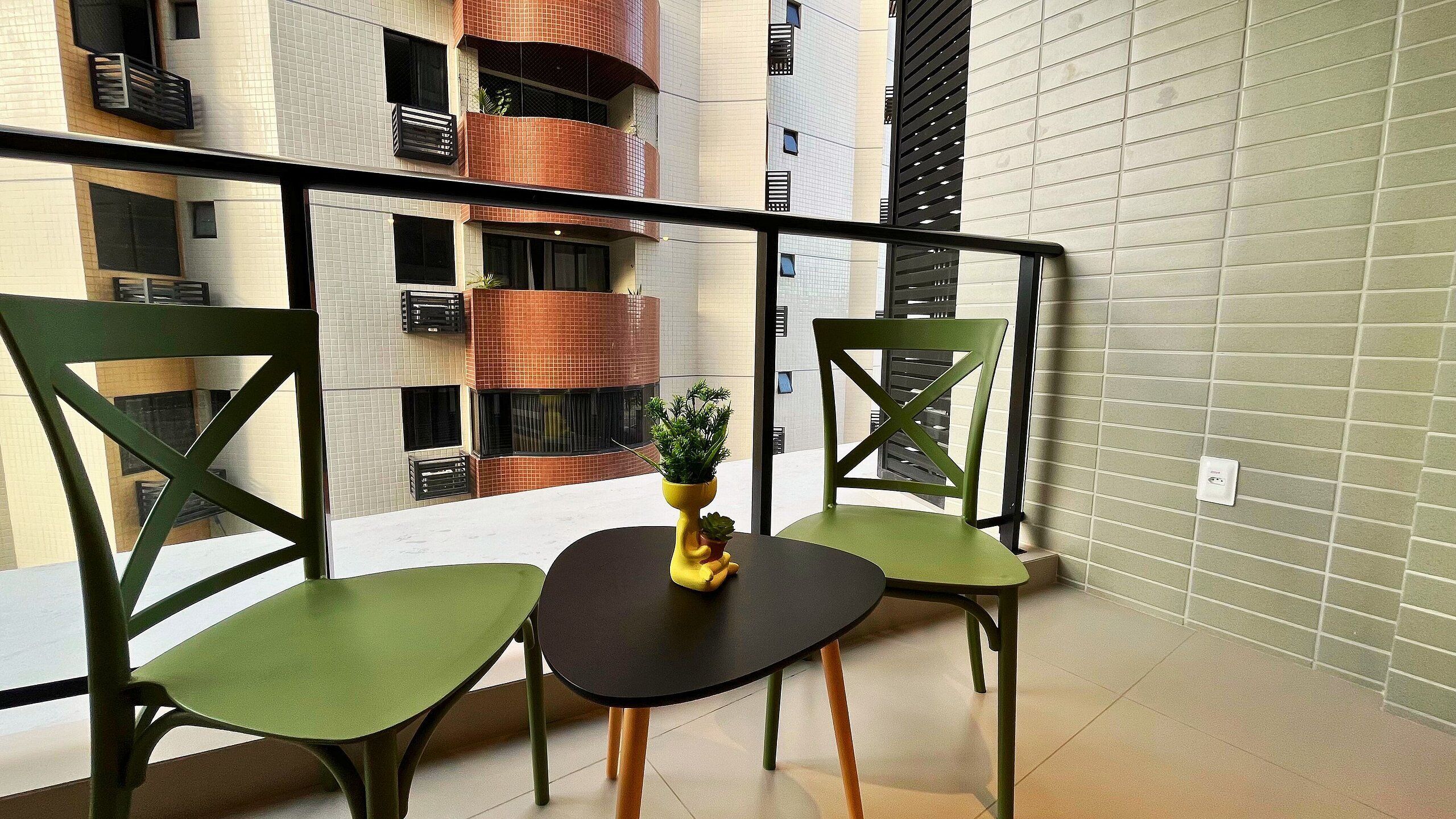 Varanda com duas cadeiras verdes, uma mesa pequena, planta em vaso e vista de um edifício com várias varandas.