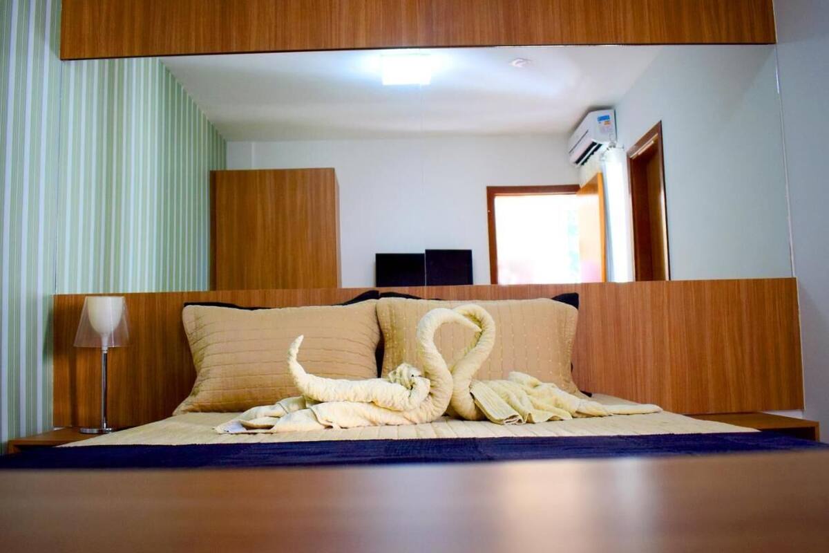 Quarto de hotel com cama, cabeceira de madeira, mesa de cabeceira com abajur e ar condicionado fixado na parede.